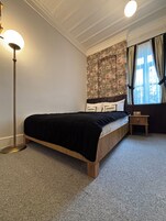 Quarto com cama, mesa de cabeceira, abajur, janela com cortinas e papel de parede com padrão floral.