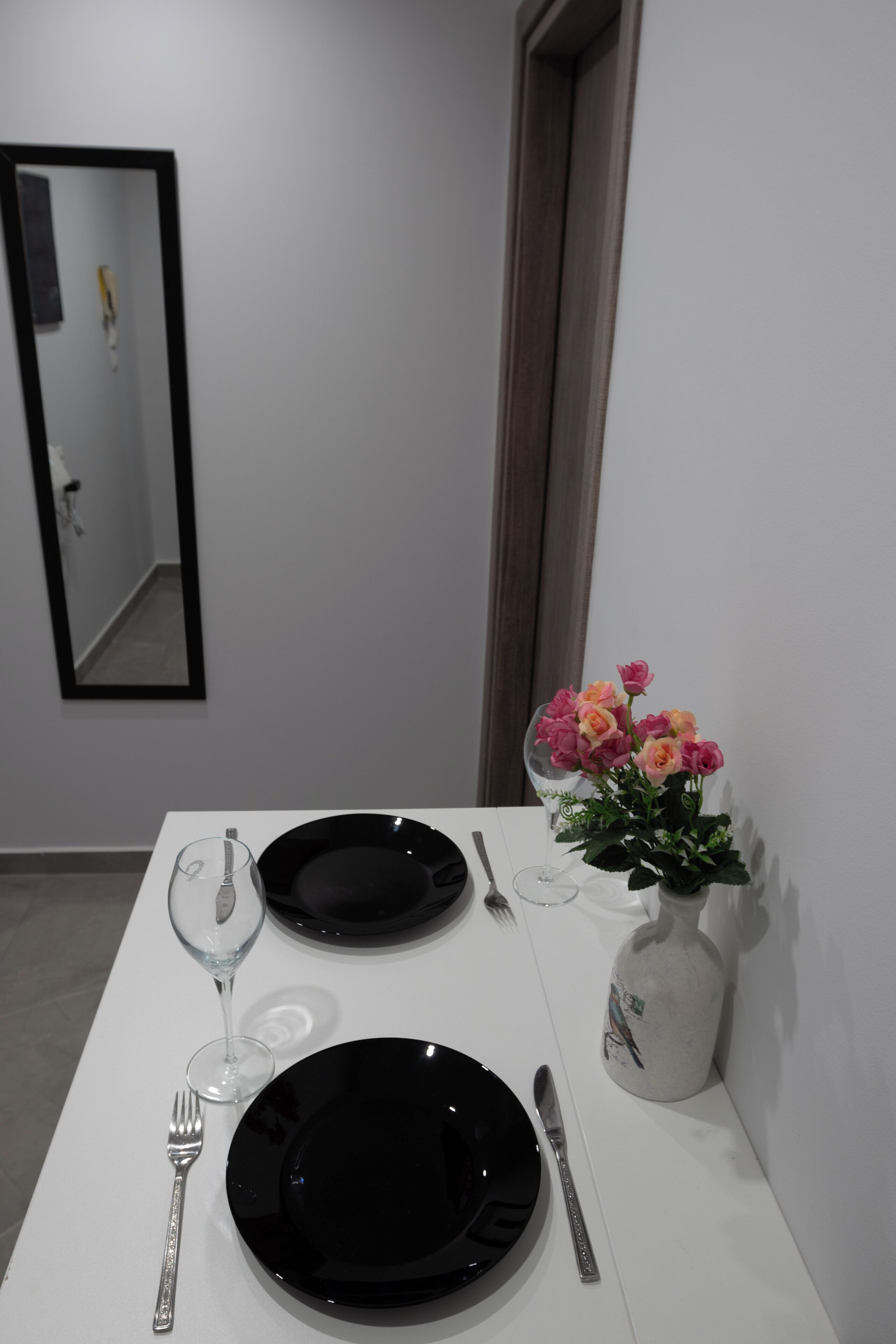 Mesa de jantar posta com pratos pretos, talheres e um vaso com flores.