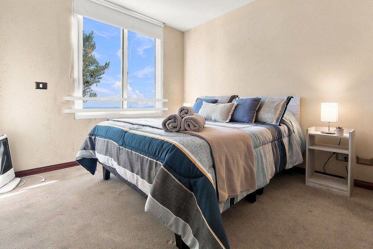 Quarto com uma janela grande, cama com colcha listrada, abajur e pequena mesa com planta.