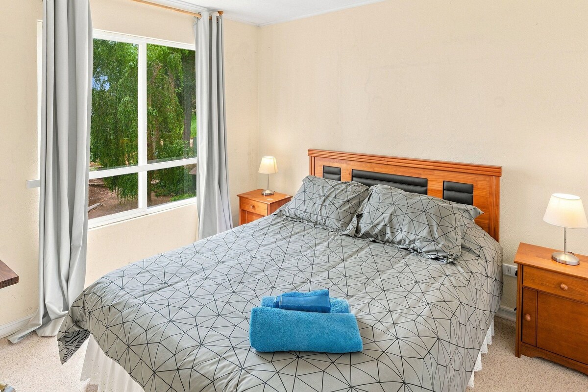 Quarto com cama, duas mesas de cabeceira, janela com cortinas e vista para o verde lá fora.