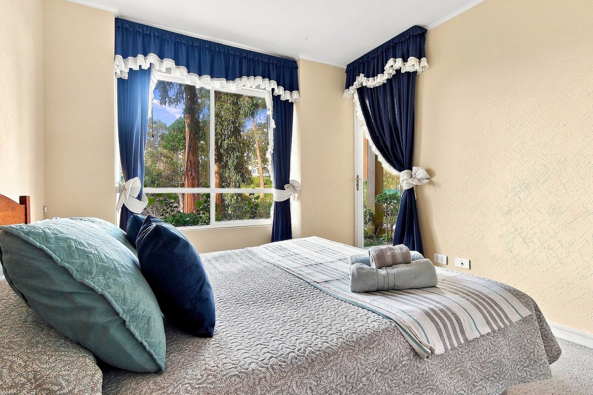 Quarto com cama, duas almofadas, janela com cortinas e vista para o verde lá fora.