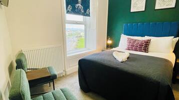 Quarto de hotel com uma cama, uma cadeira, um radiador, uma janela com vista e duas imagens emolduradas na parede.