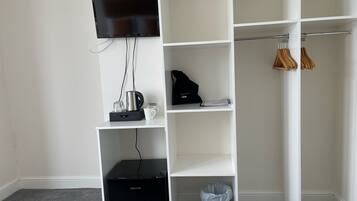 Um quarto com uma TV fixada na parede, um aquecedor elétrico preto, um cesto de lixo e um cabideiro com um casaco.