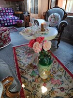 Uma sala de estar aconchegante com uma mesa redonda posta para refeições, uma poltrona florida e um vaso decorativo com rosas.