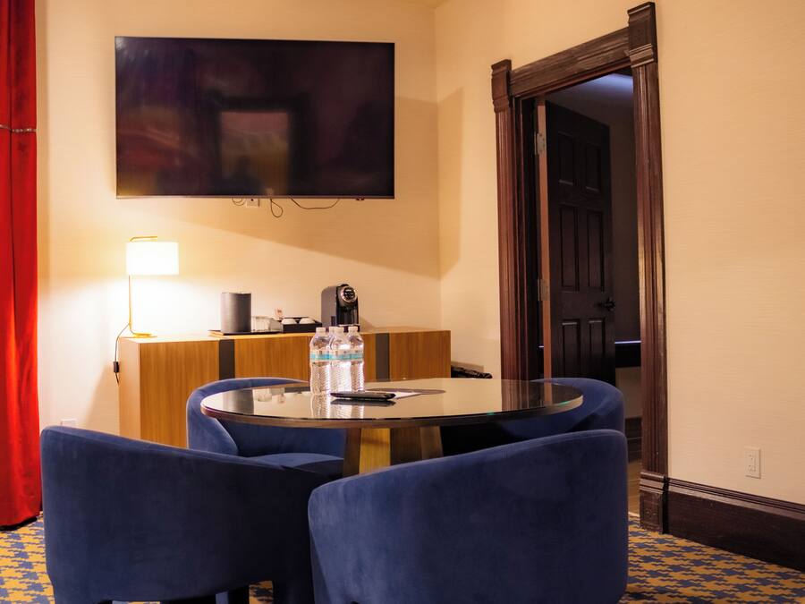 Quarto de hotel com mesa de vidro redonda, cadeiras estofadas em azul, televisão de tela plana na parede, escrivaninha de madeira com abajur e cortina vermelha.