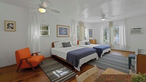 Quarto de hotel com duas camas, uma cadeira, uma escrivaninha e um ventilador de teto.