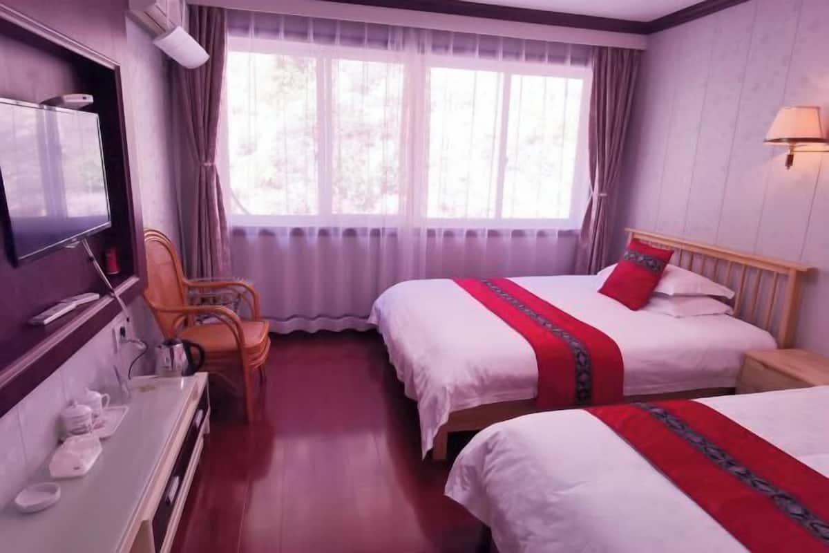 Quarto de hotel com duas camas, televisão, cadeira, mesa de cabeceira e janela com cortinas.