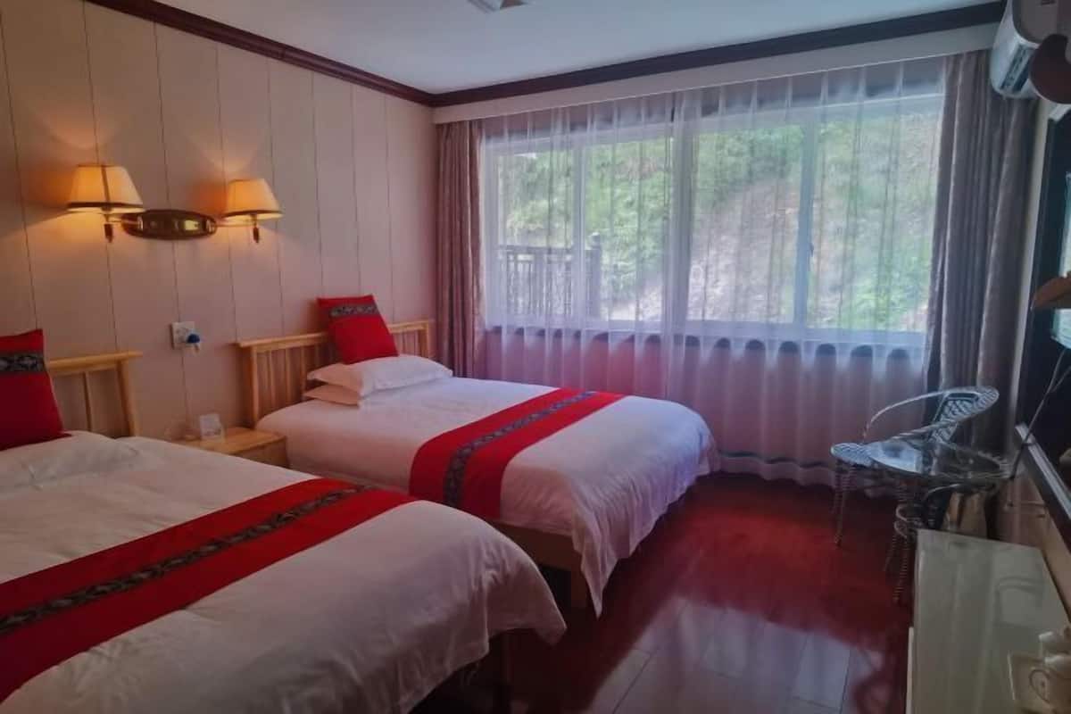 Quarto de hotel com duas camas, uma cadeira, uma mesinha de cabeceira e uma janela grande com cortinas.