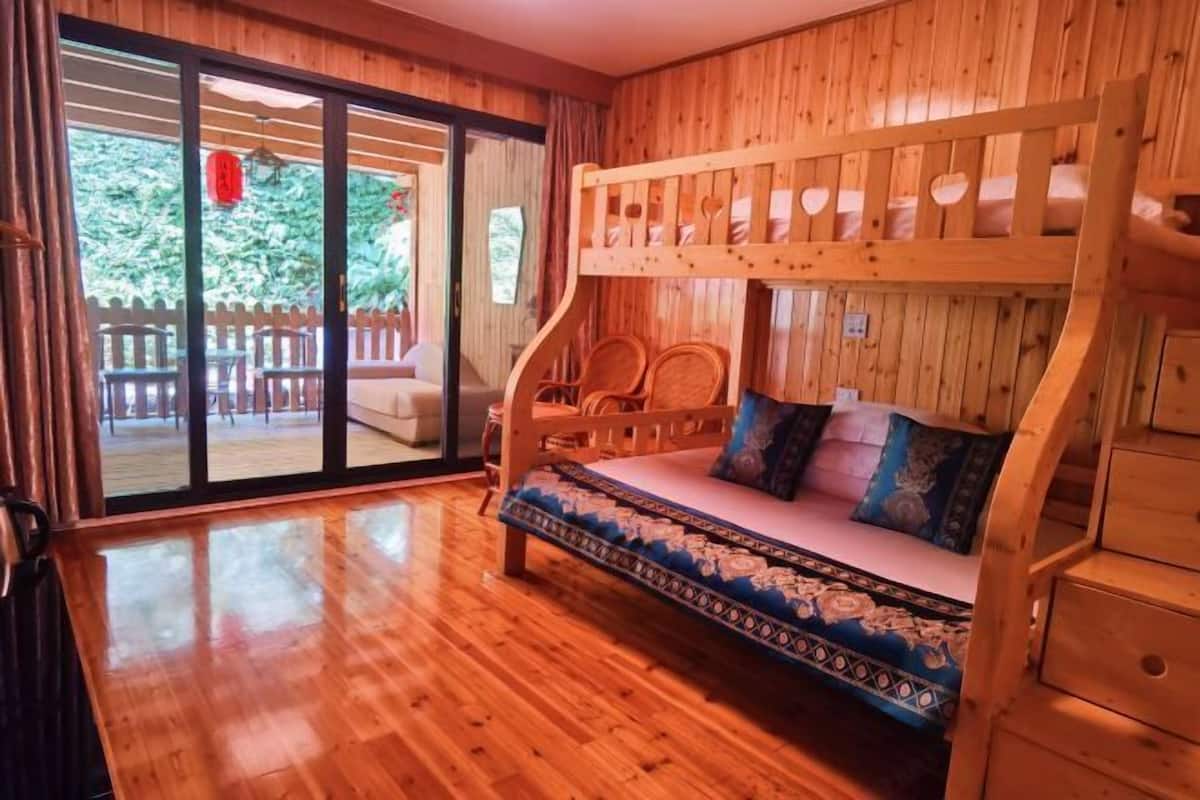 Quarto de madeira com beliche, sofá com almofadas e uma porta de correr de vidro que dá para uma área externa com cadeiras.