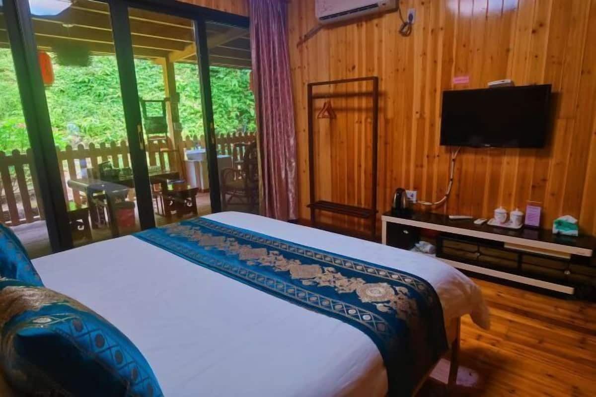Quarto de madeira com uma cama, uma televisão e vista para o verde lá fora.