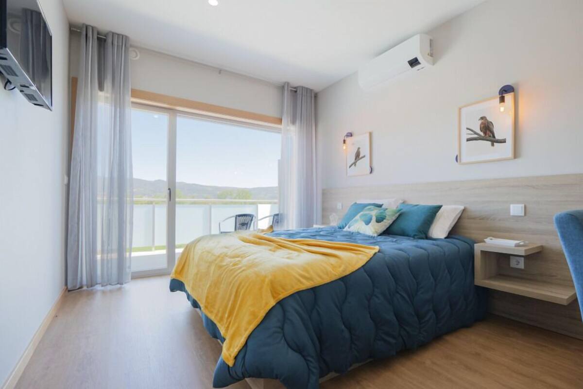 Quarto com cama, mesa de cabeceira, cadeira e janela com cortinas.