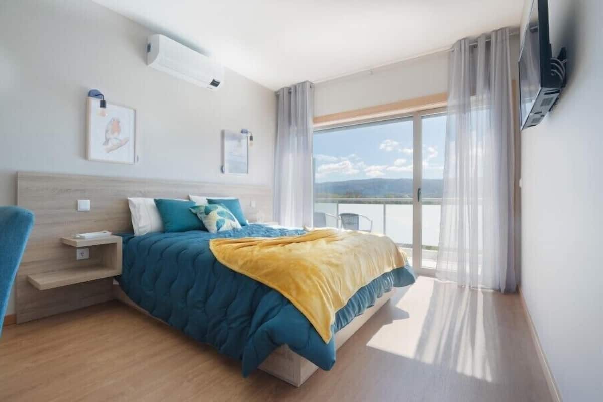 Quarto de hotel moderno com uma cama grande, uma mesa de cabeceira, televisão montada e uma janela com vista para a paisagem.