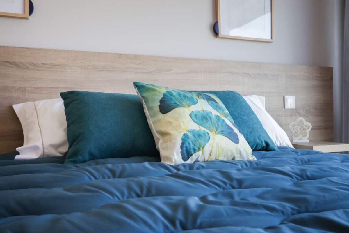 Cama bem arrumada com edredom azul e duas almofadas, uma estampada com flores e outra lisa.