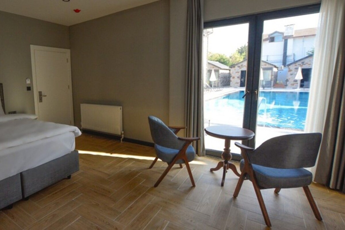 Quarto de hotel com cama, duas cadeiras, uma mesinha, um quadro e vista para a piscina.