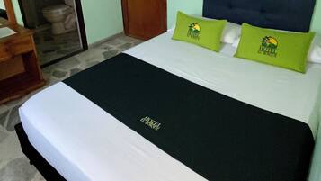 Quarto de hotel com uma cama, duas almofadas, uma porta de madeira e um banheiro visível através de uma porta aberta.