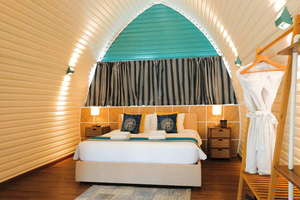 Quarto com cama, cabeceira de madeira, mesa de cabeceira, roupão pendurado e parede com cortina.