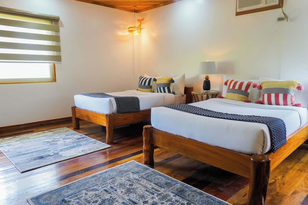 Quarto de hotel com duas camas de madeira, uma janela com persianas, um candeeiro fixado na parede e uma pequena mesa com abajur.