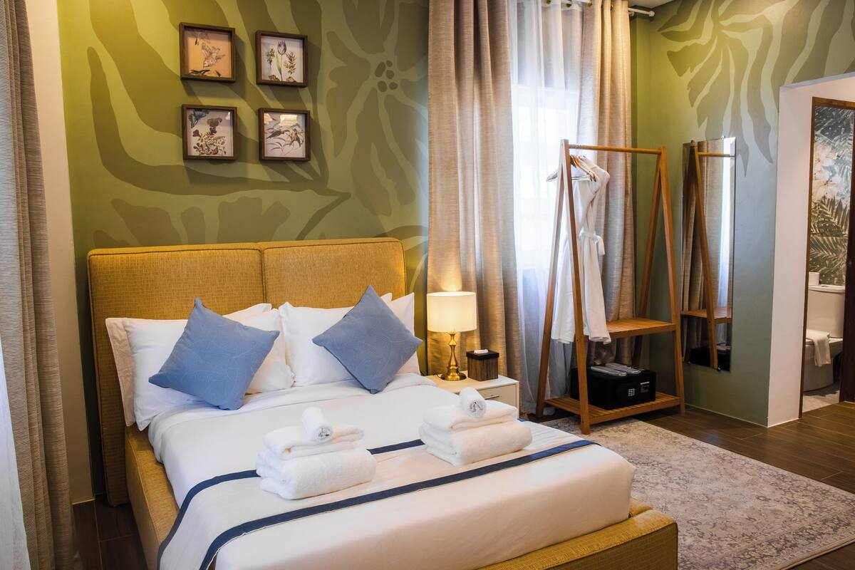 Cama bem arrumada com lençóis brancos e almofadas azuis, uma luminária de cabeceira e um quadro na parede.
