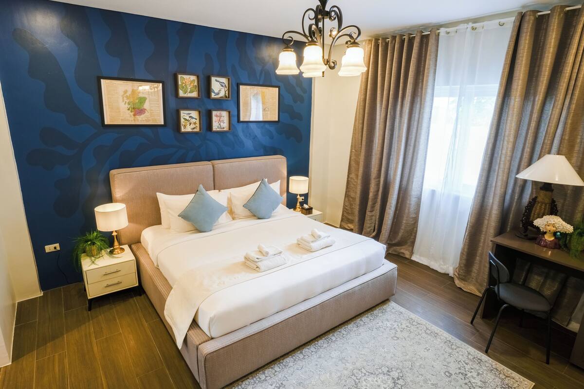 Quarto bem organizado com uma cama grande, mesas de cabeceira, uma escrivaninha com cadeira e uma janela com cortinas.
