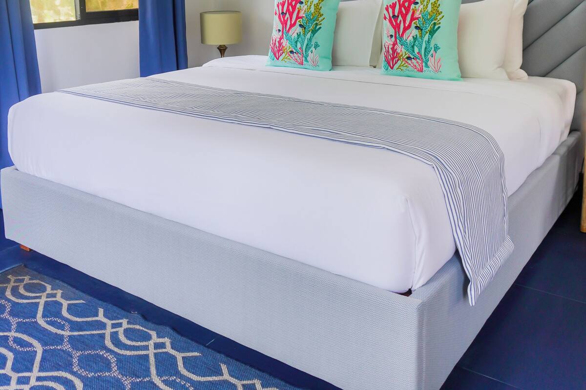 Quarto de hotel com uma cama grande, uma janela com cortinas azuis, um quadro de flamingos e um tapete estampado.