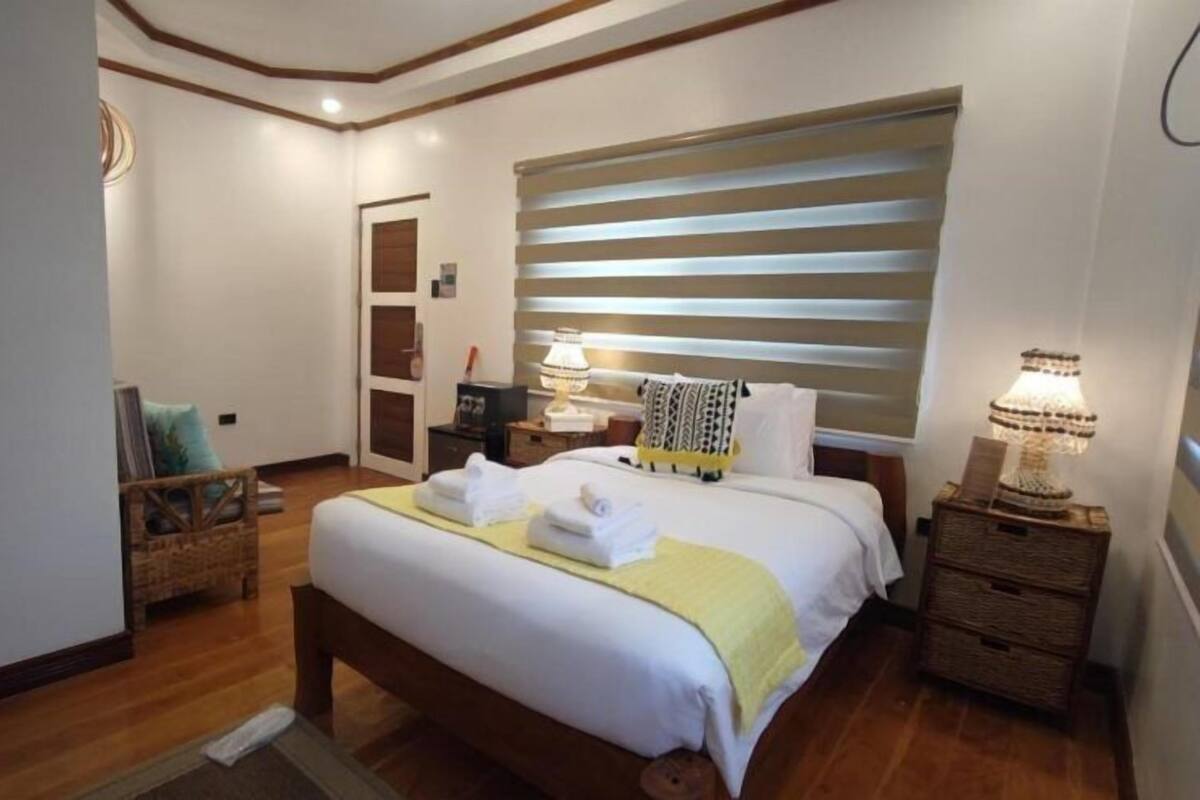 Quarto com uma cama grande, duas mesas de cabeceira, um criado-mudo, uma luminária na parede e uma janela com persianas.