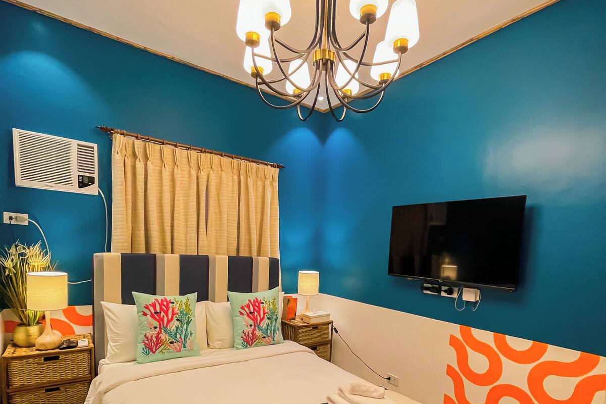 Quarto de hotel moderno com uma cama grande, um lustre, uma TV de tela plana e uma parede azul de destaque.