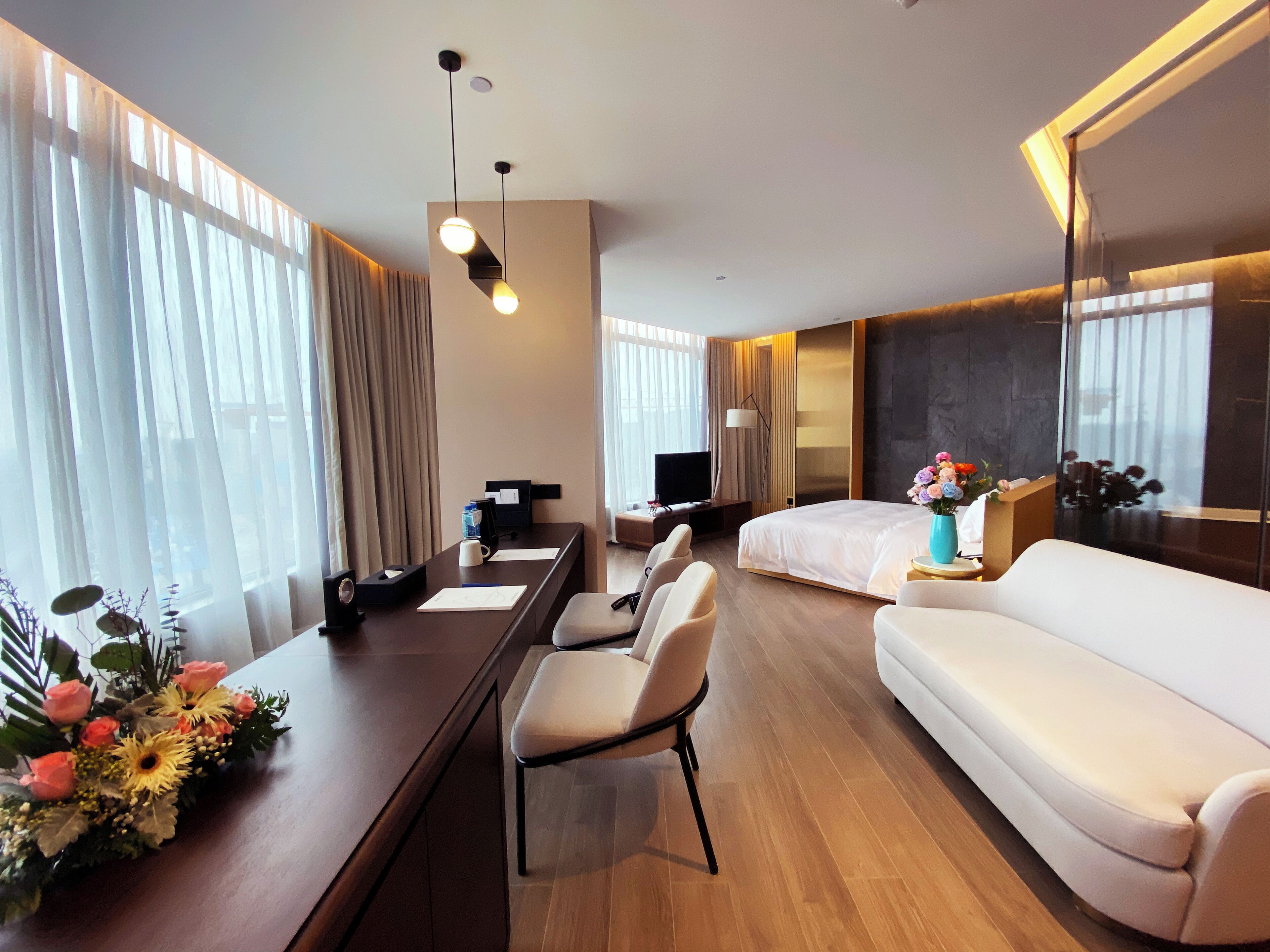 Quarto de hotel moderno com cama, sofá, escrivaninha com cadeiras, televisão e uma janela grande com cortinas.
