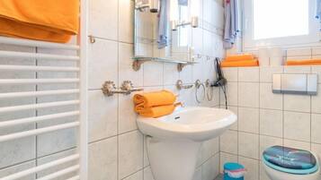 Um banheiro com pia branca, espelho e vaso sanitário. Há um porta-toalhas com toalhas laranja, uma lixeira azul e uma janela com cortinas.