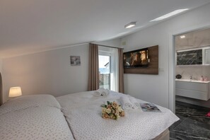 Quarto de hotel com cama, um buquê de flores, uma revista, uma televisão e banheiro com piso de mármore.