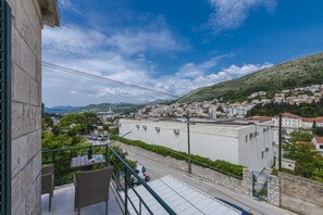 Varanda com cadeiras de vime e mesa, com vista para uma cidade em encosta com edifícios e vegetação.
