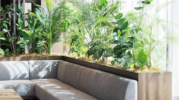 Uma sala de estar moderna com um sofá seccional cinza, uma mesa de centro de madeira e um arranjo de plantas em vasos grandes, tudo em frente a uma janela com cortinas leves.