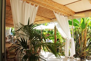 Área externa coberta com teto de bambu, cortinas brancas e plantas tropicais exuberantes.
