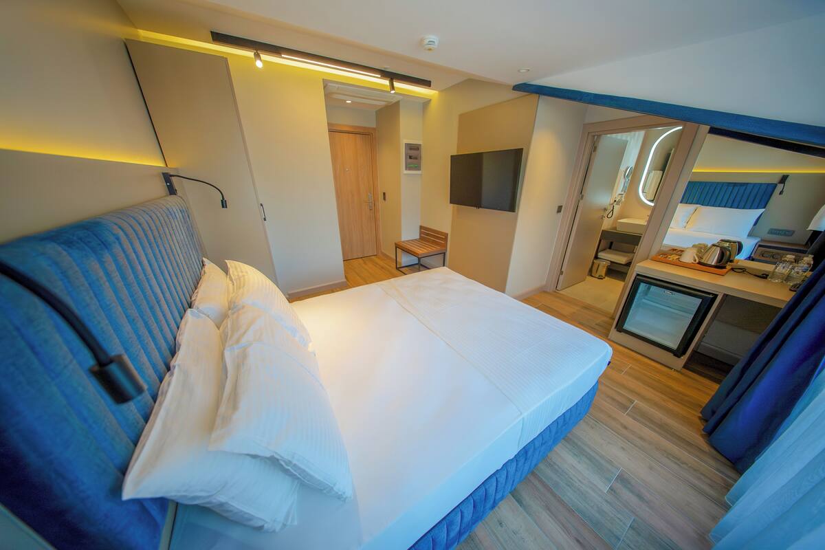 Quarto de hotel moderno com cama, escrivaninha, TV e uma pequena cozinha equipada.