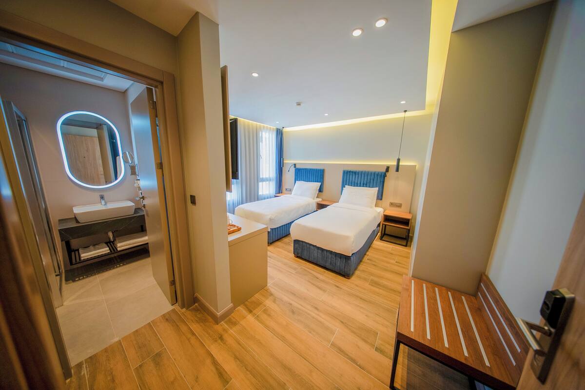Quarto de hotel com banheiro, duas camas, mesa de cabeceira e espelho iluminado.