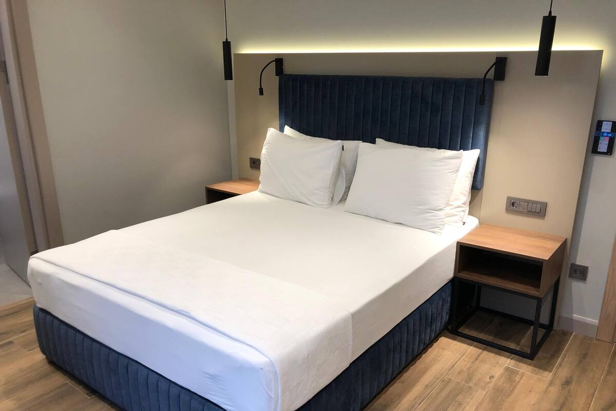 Quarto de hotel moderno com uma cama grande, mesas de cabeceira de madeira e uma cabeceira com iluminação integrada.