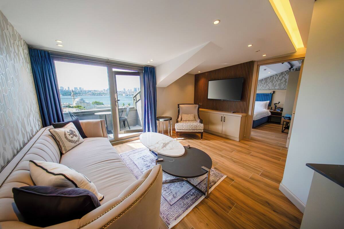 Quarto de hotel moderno com sofá, uma mesinha, TV e vista para a cidade.