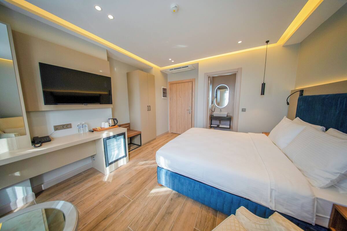 Quarto de hotel moderno com uma cama grande, uma TV de tela plana na parede, uma pequena escrivaninha com cadeira e banheiro com espelho e pia.