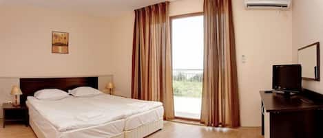 Quarto de hotel com cama, escrivaninha com televisão, mesa de cabeceira com abajur e janela com cortinas.