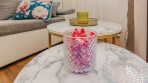 Recipiente de vidro com objetos coloridos sobre uma mesa de mármore, um sofá com almofadas florais e uma vela verde ao fundo.