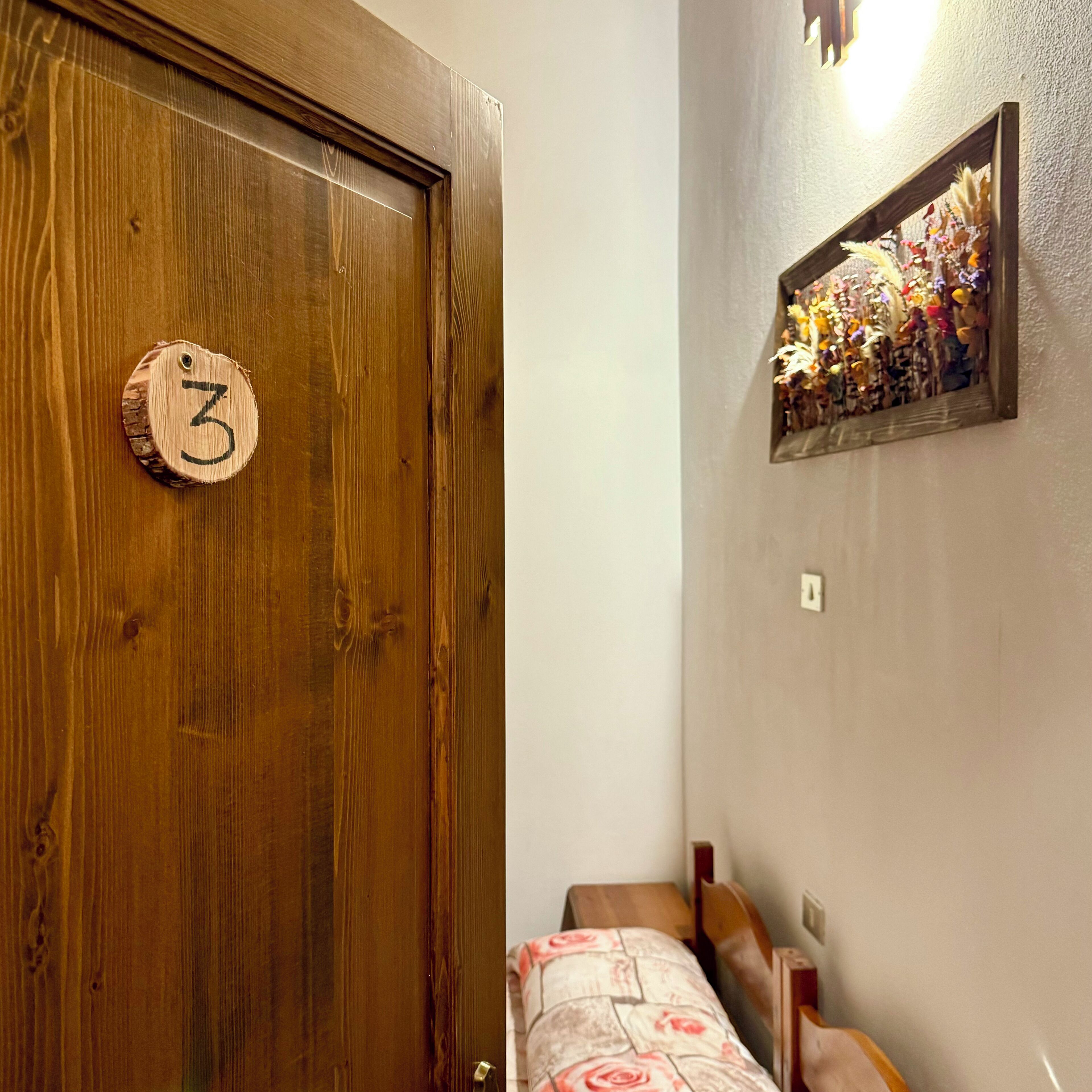 Uma porta de madeira com uma placa de número, uma cama com colcha floral e um objeto decorativo na parede.