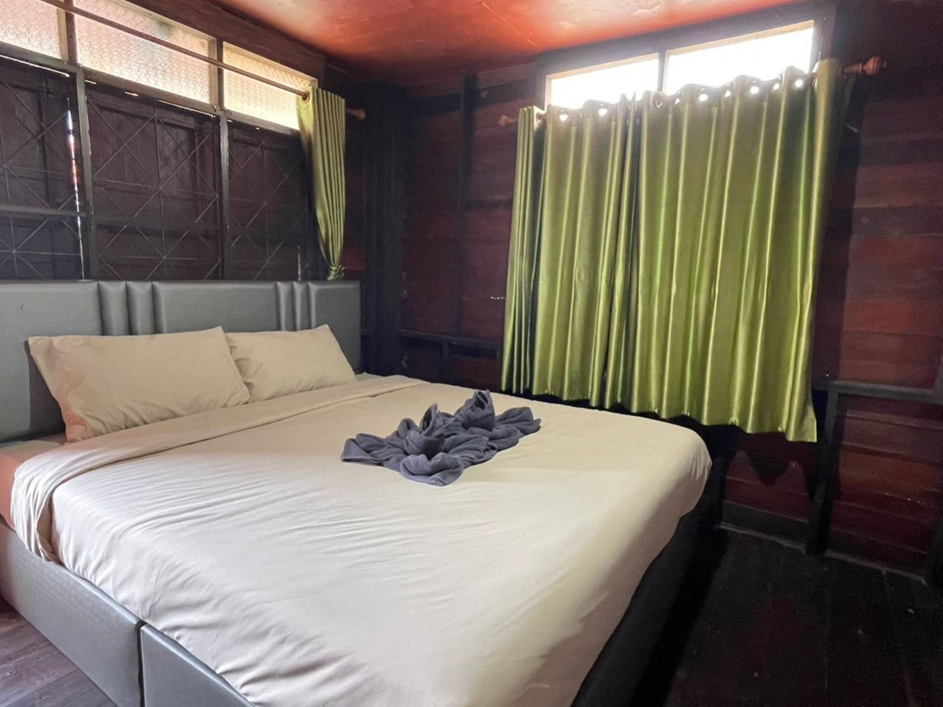 Cama bem arrumada com colcha e almofadas bege, janela com cortinas verdes e cabeceira de madeira.