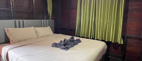 Cama bem arrumada com colcha e almofadas bege, janela com cortinas verdes e cabeceira de madeira.