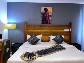 Quarto de hotel com cabeceira de madeira, pintura de uma pessoa à beira-mar, luminária de mesa e cama bem arrumada com almofadas estampadas e capa decorativa para a cabeceira.