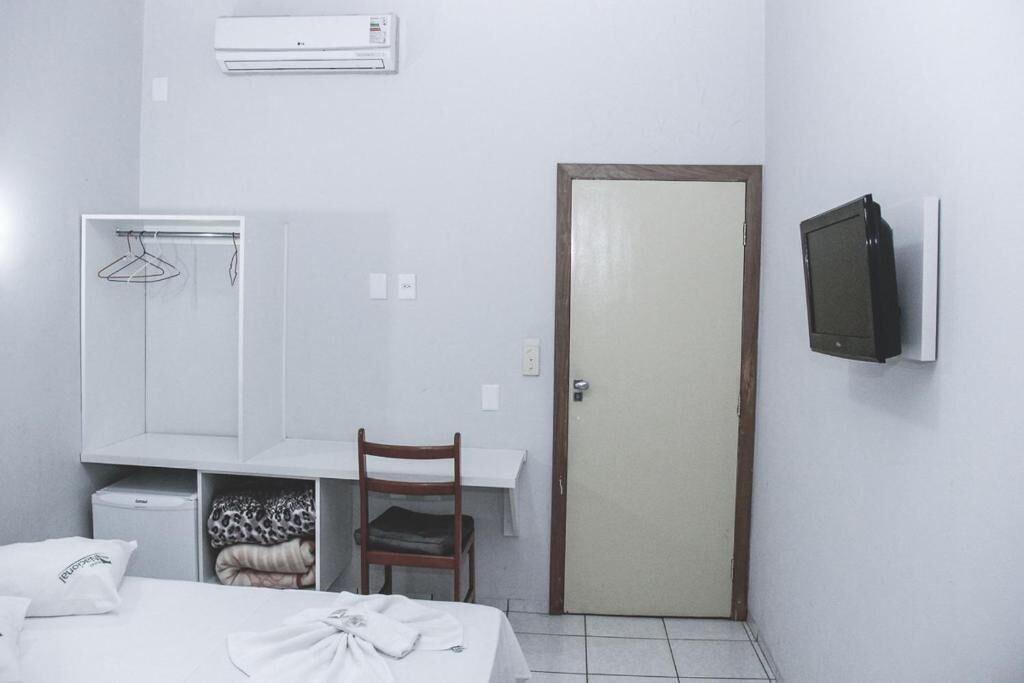 Quarto pequeno e limpo, com cama, cadeira, guarda-roupa, televisão e porta.