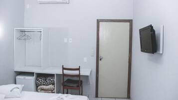 Quarto pequeno e limpo, com cama, cadeira, guarda-roupa, televisão e porta.