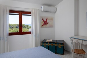 Quarto com cama, janela com cortinas, ar condicionado fixado na parede, escrivaninha de madeira, cadeira e um baú azul com um livro.