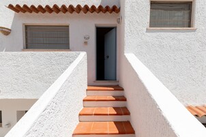 Um edifício branco com porta azul e degraus de barro que levam a uma janela.