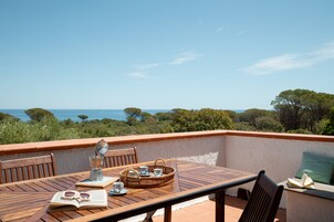 Uma mesa de madeira com um livro, óculos de sol e uma cafeteira numa varanda com vista para o mar e árvores.