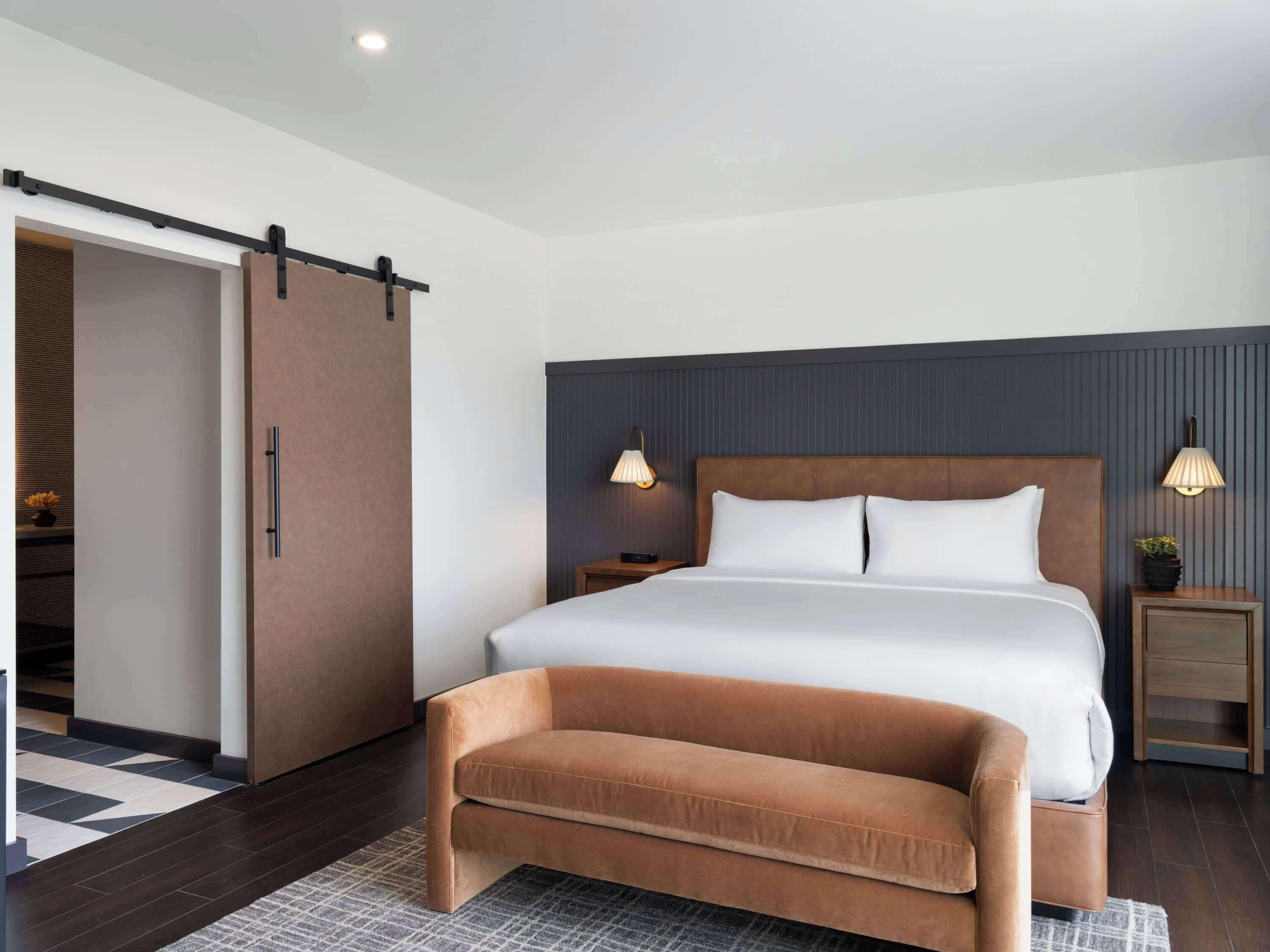 Quarto moderno com uma cama grande, um sofá marrom, uma porta deslizante e duas luminárias de cabeceira.
