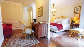 Quarto de hotel com secretária de madeira, poltrona vermelha, cama branca com detalhes dourados e um candelabro.
